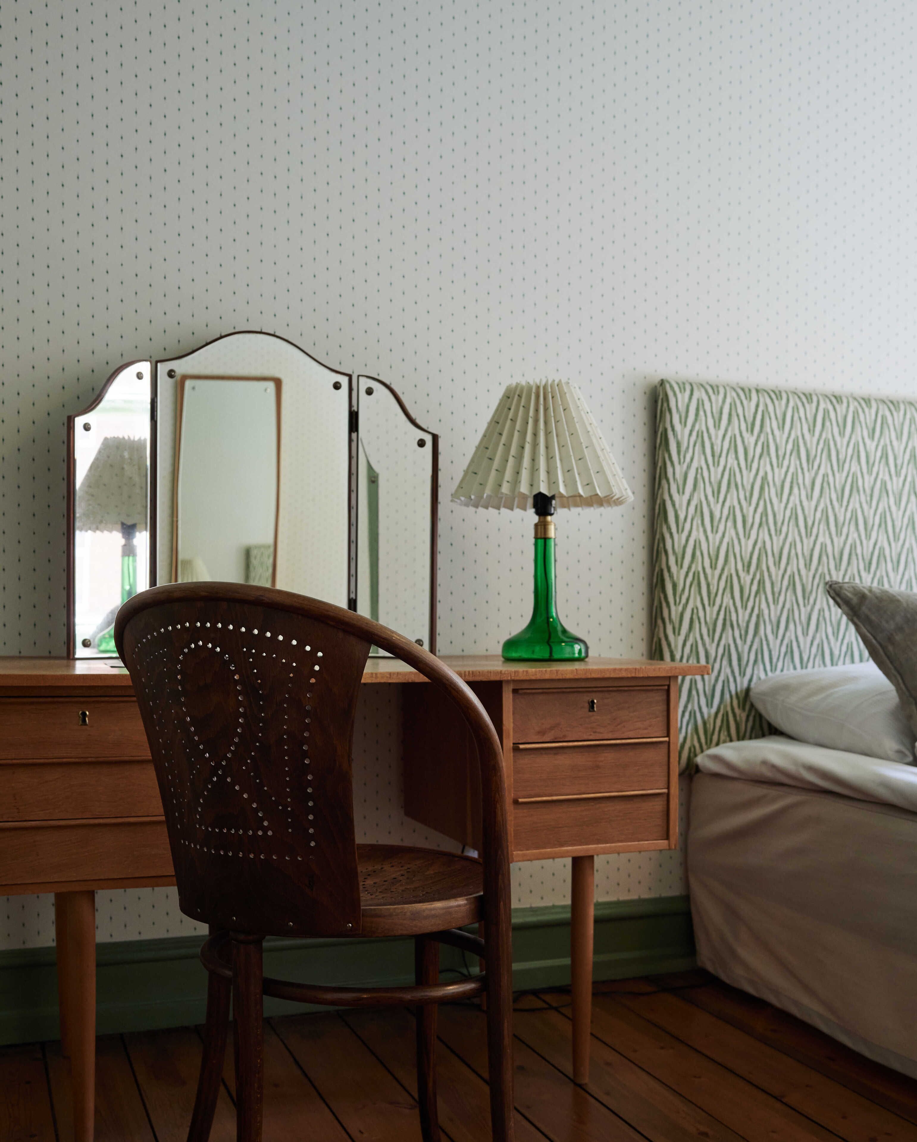 Uma escrivaninha de madeira com um espelho, uma luminária verde e uma cama com cabeceira estampada.