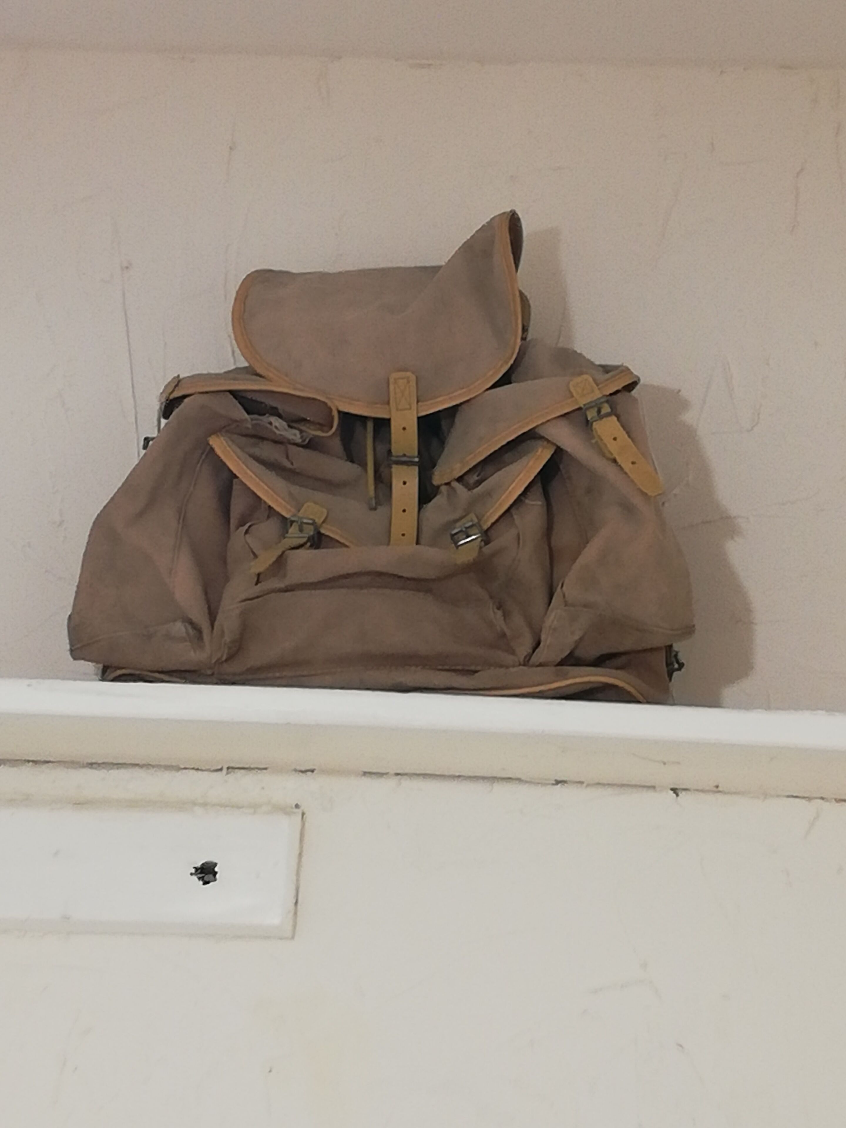 Uma mochila marrom com várias alças e fivelas, colocada sobre uma prateleira branca em frente a uma parede lisa.