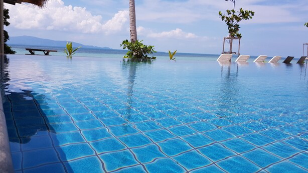 Uma piscina azul infinita com vista para o oceano e montanhas, cercada por palmeiras e cadeiras de lounge.