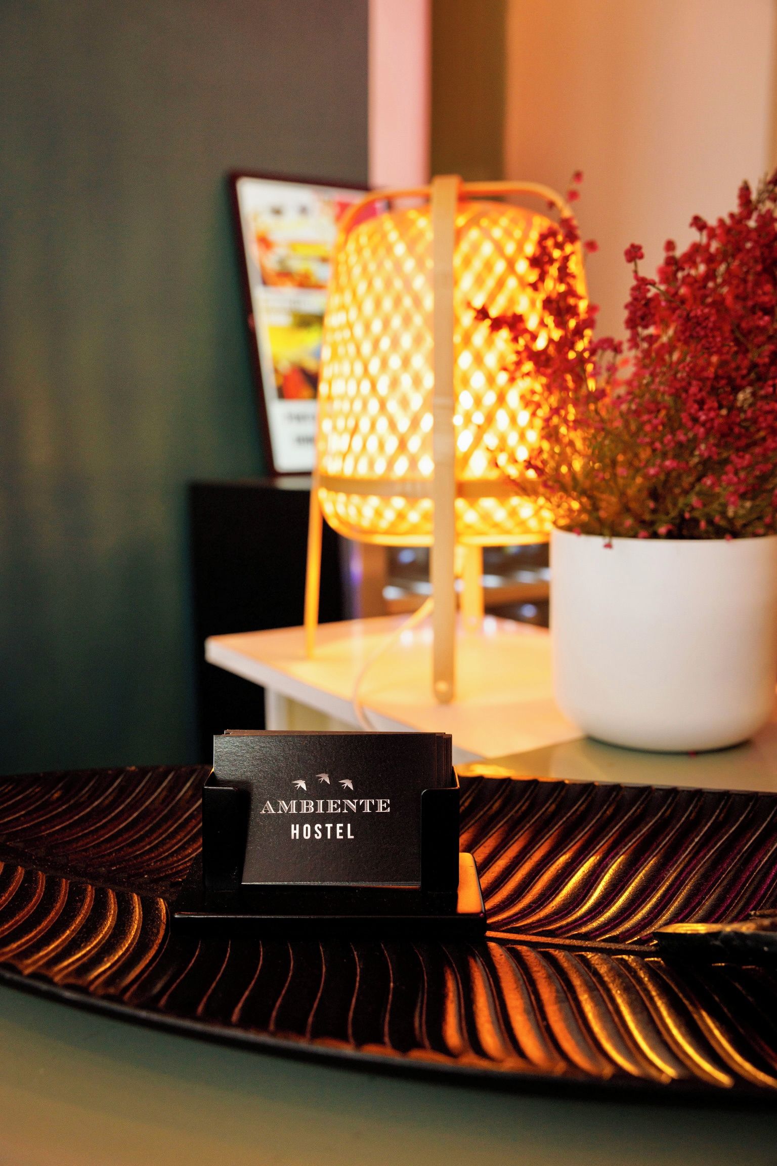 Placa com a inscrição 'AMBIENTE HOSTEL', com um abajur decorativo e uma planta em vaso ao fundo.