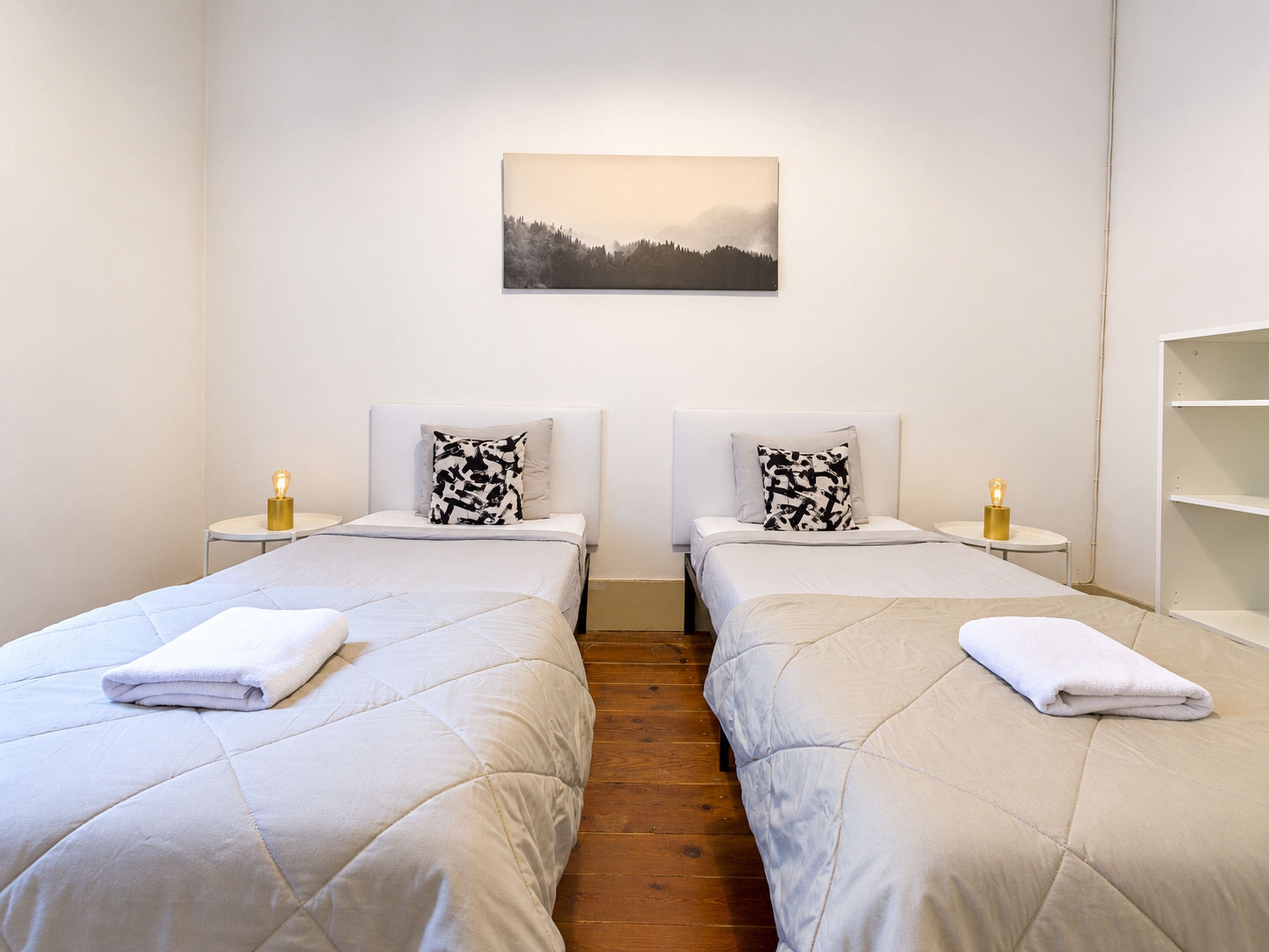Quarto de hotel com duas camas, uma mesa de cabeceira com abajur, um quadro na parede e chão de madeira.