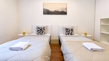 Quarto de hotel com duas camas, uma mesa de cabeceira com abajur, um quadro na parede e chão de madeira.