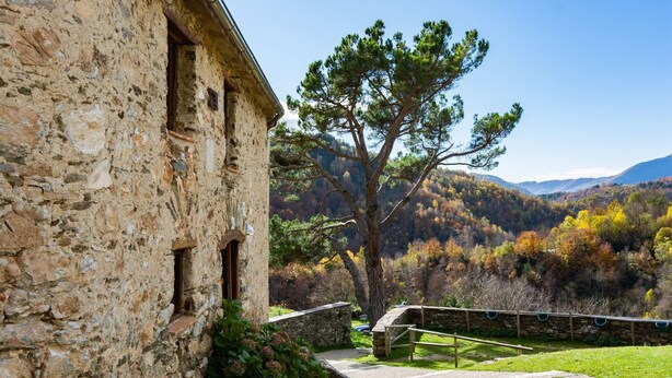Um edifício de pedra com aspecto rústico, uma grande árvore em frente e vista para uma paisagem montanhosa.