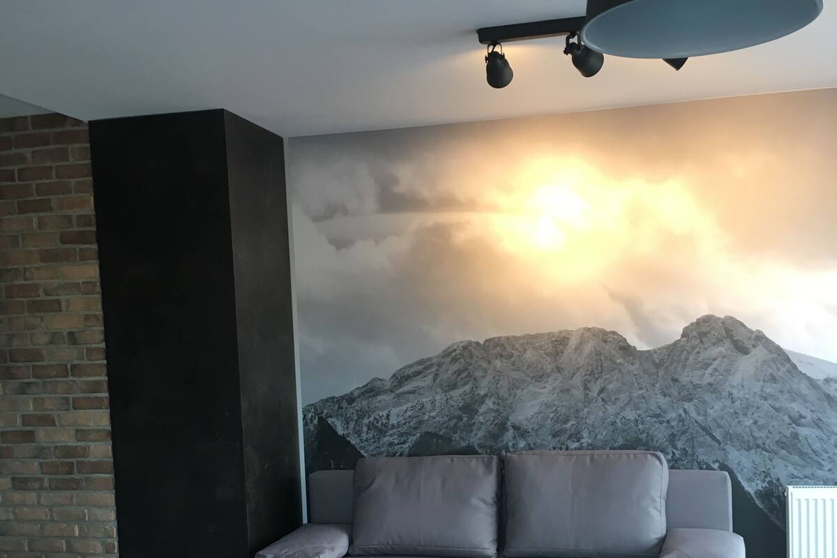 Uma sala de estar moderna com um sofá cinzento, uma luminária preta e um mural com tema de montanhas na parede.