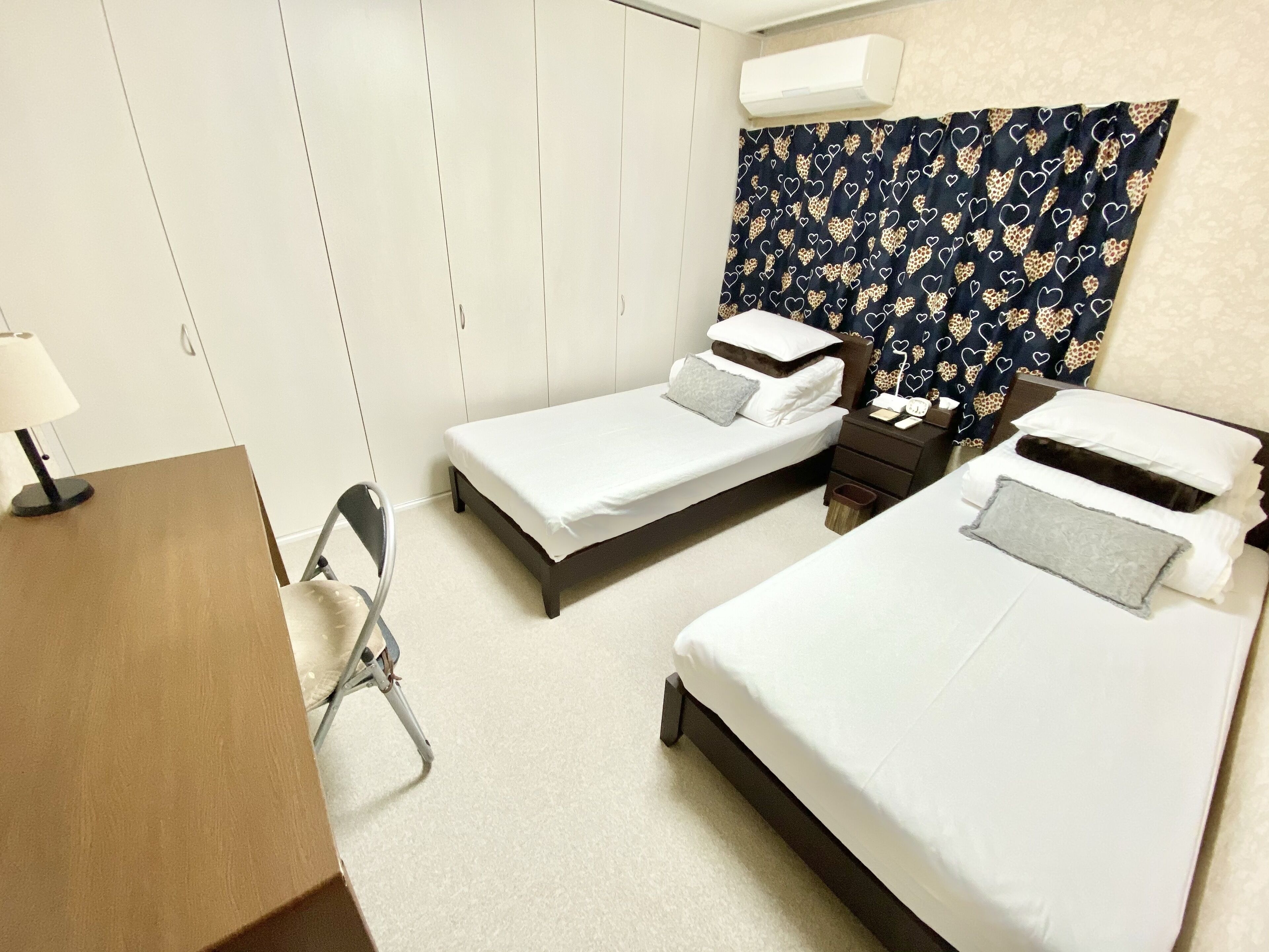 Quarto de hotel com duas camas de solteiro, uma escrivaninha com luminária, uma cadeira, um guarda-roupa e cortina estampada.