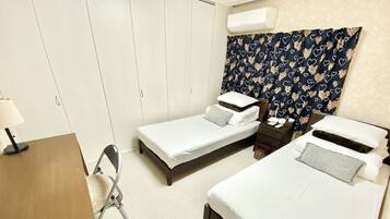 Quarto de hotel com duas camas de solteiro, uma escrivaninha com luminária, uma cadeira, um guarda-roupa e cortina estampada.