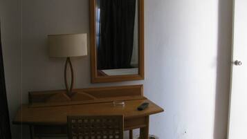 Quarto de hotel com escrivaninha de madeira, luminária, espelho, cadeira e lixeira.