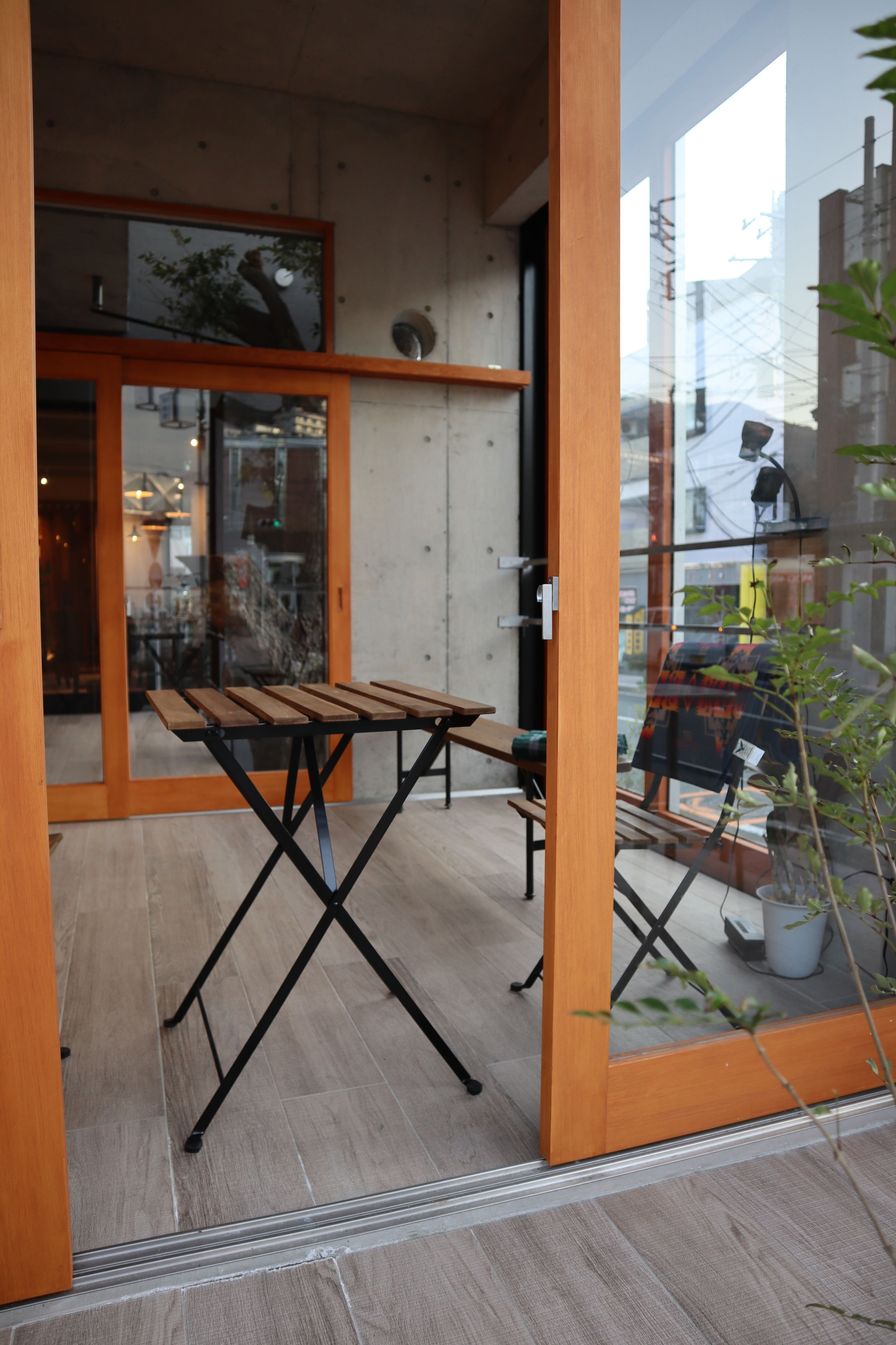 Uma área de estar externa com mesa e bancos de madeira, porta de vidro que dá para o interior e vista da rua e prédios pelo vidro.
