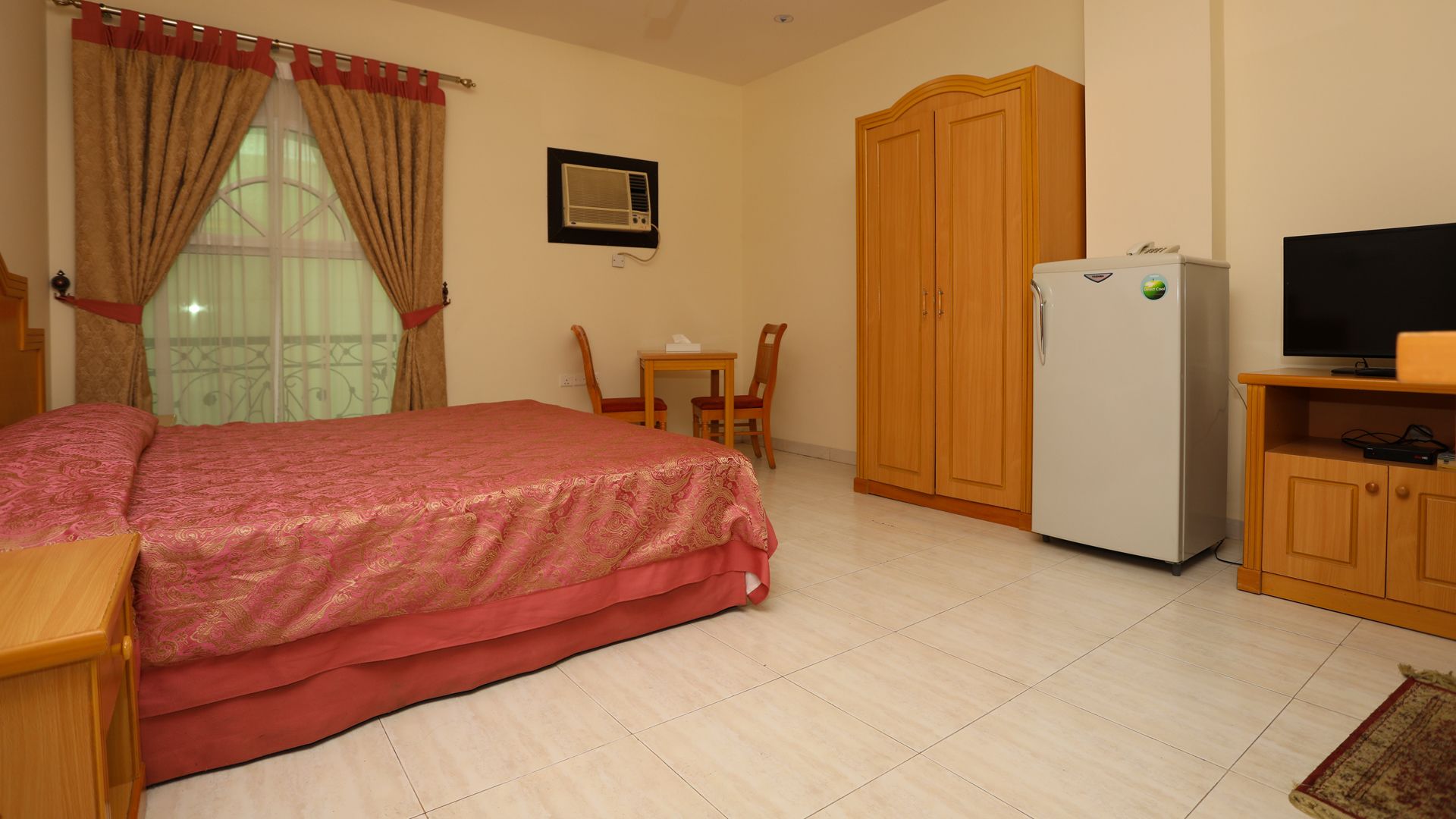 Quarto de hotel com cama, escrivaninha, cadeira, guarda-roupa, televisão e frigorífico.
