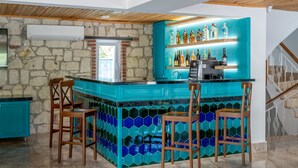 Uma área de bar com bancada turquesa e banquetas, cercada por paredes de pedra e teto de madeira.