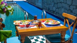 Uma mesa de jantar com pratos de comida, um copo de chá e uma bandeja de metal, ao fundo uma parede de pedra e uma piscina.
