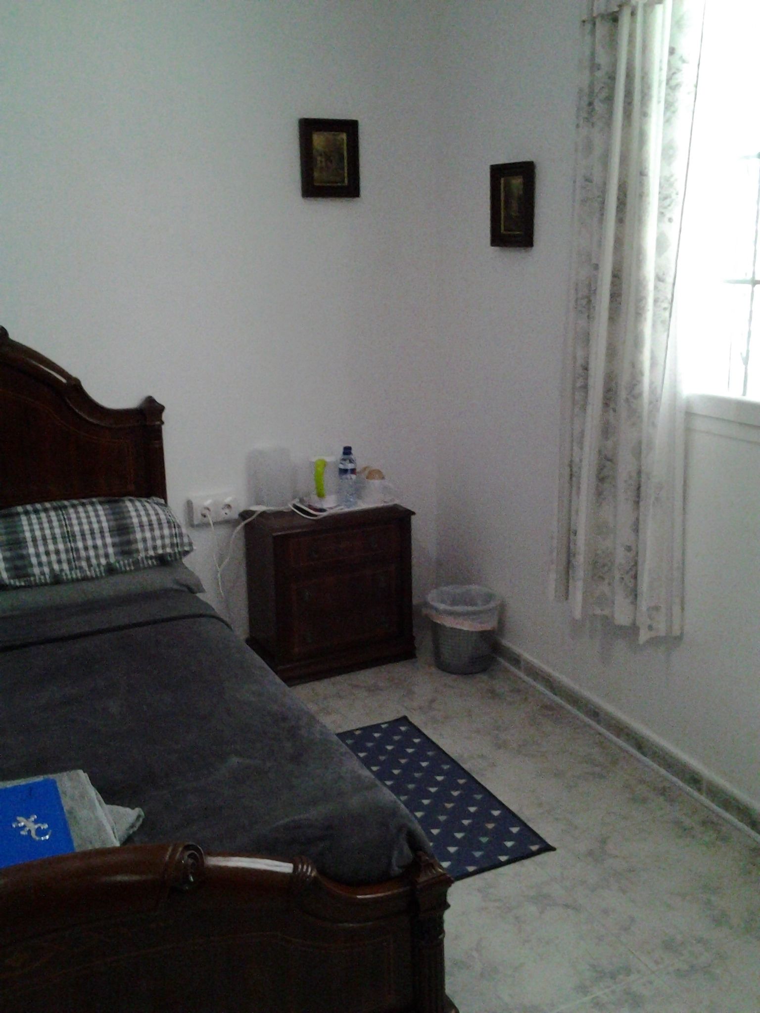 Um quarto com uma cama, uma mesa de cabeceira, uma lixeira e duas fotos emolduradas na parede.