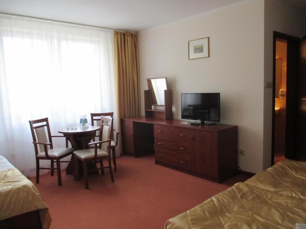 Quarto de hotel com cama, escrivaninha com televisão, cadeira e uma mesinha.
