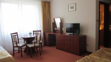 Quarto de hotel com cama, escrivaninha com televisão, cadeira e uma mesinha.