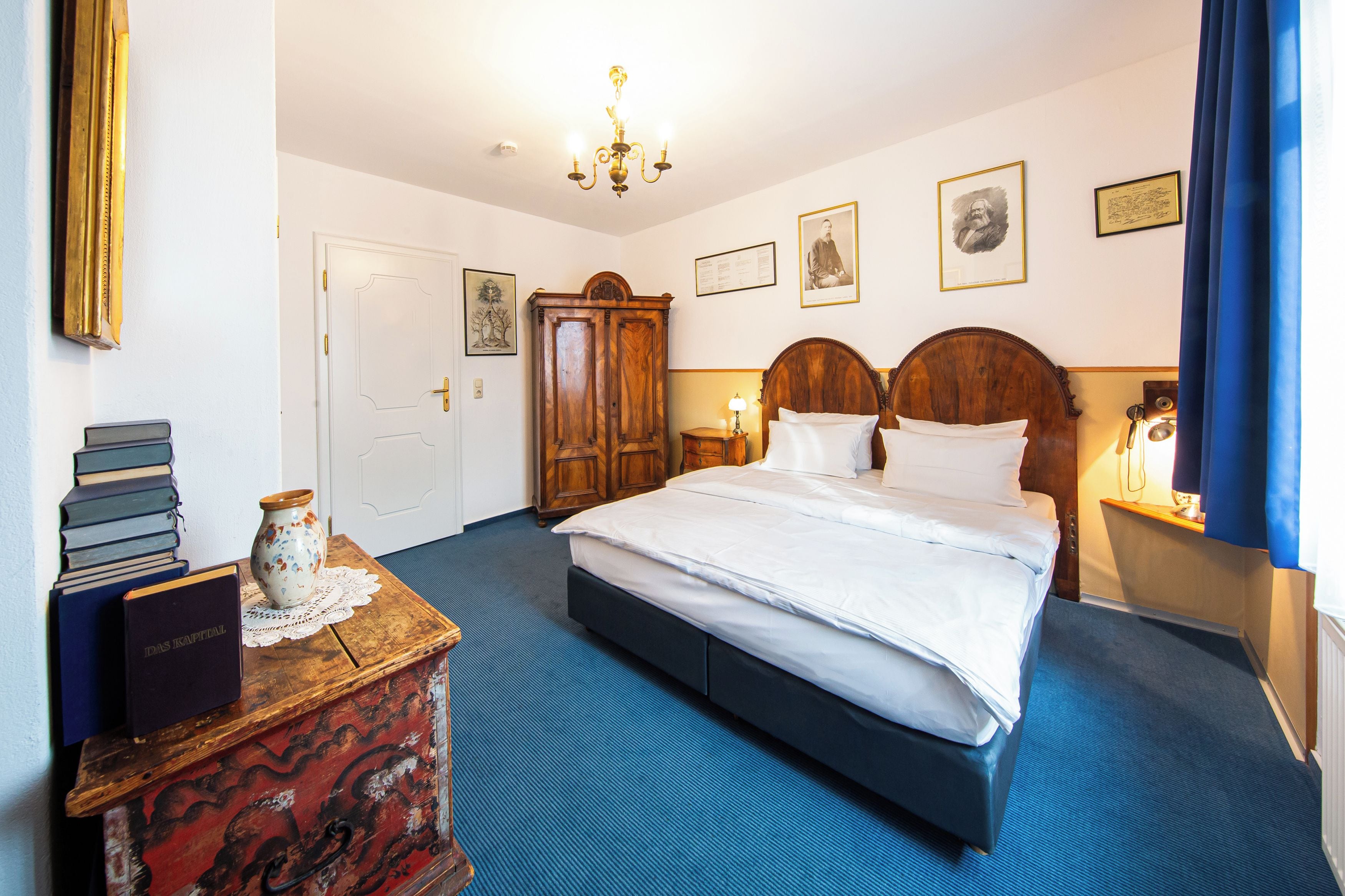 Quarto de hotel com cama grande, guarda-roupa de madeira, escrivaninha com livros e lustre.