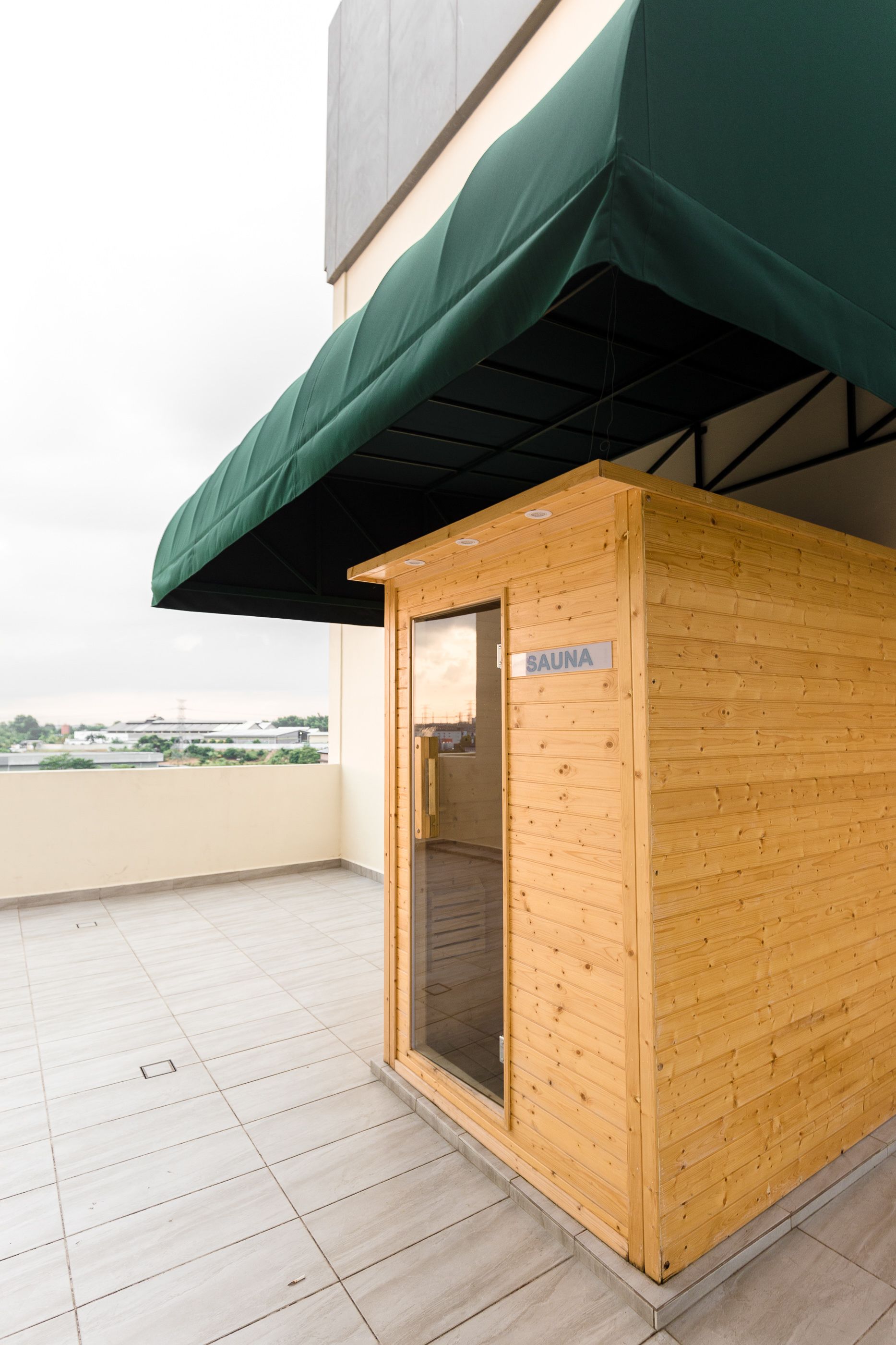 Uma sauna com exterior de madeira e uma placa com a inscrição 'SAUNA' em um terraço no telhado, com piso ladrilhado e toldo verde acima.