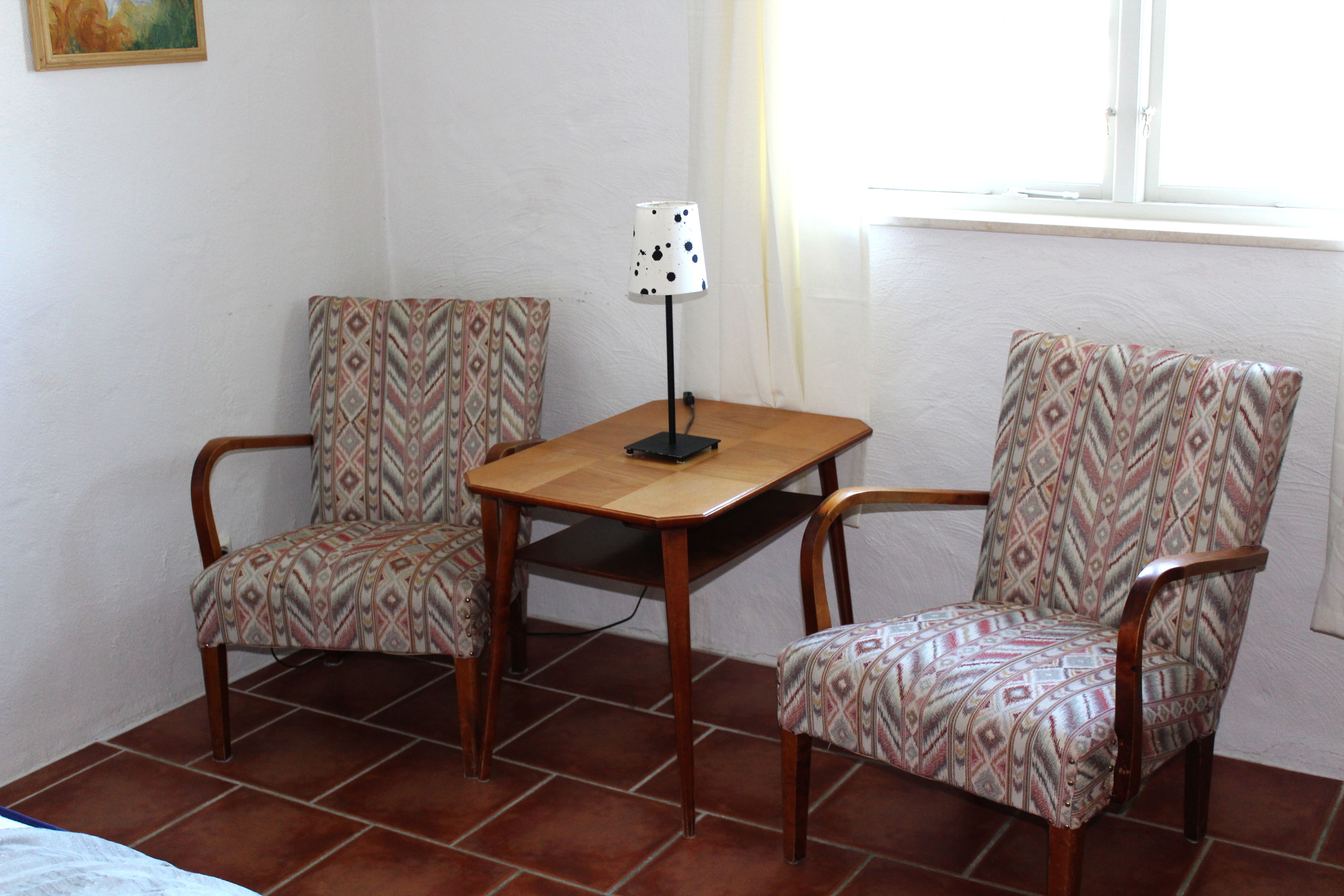 Um quarto pequeno com duas cadeiras estampadas, uma mesa de madeira e um abajur.