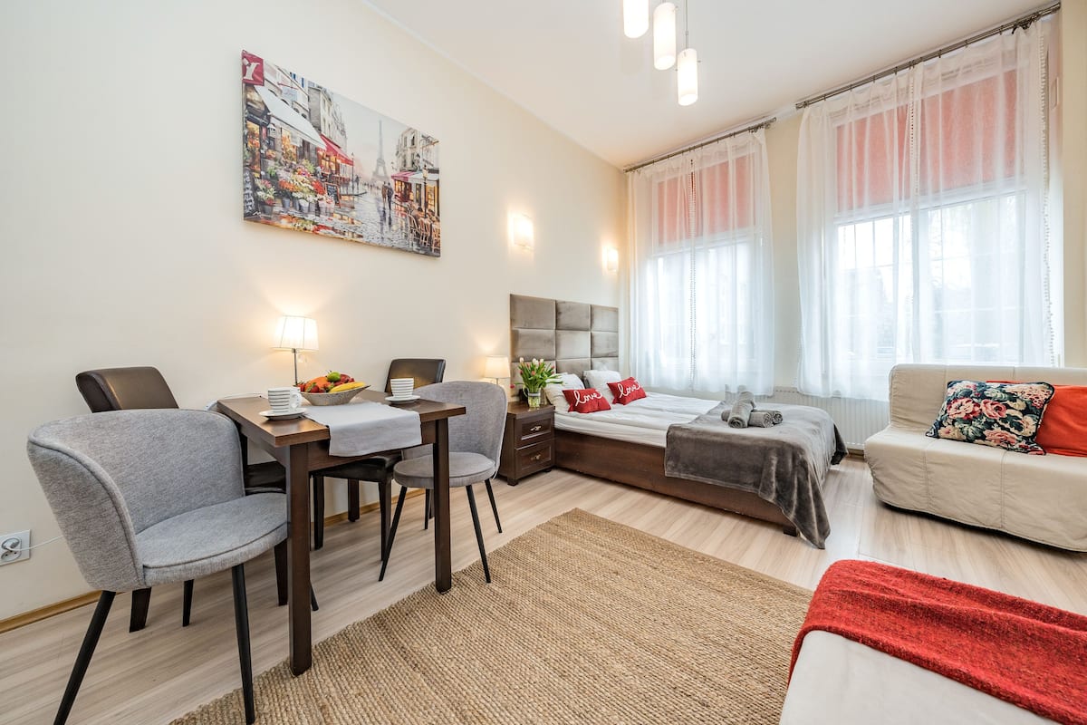 Quarto de hotel moderno com área de refeições, cama com cabeceira cinza, sofá com almofada floral e uma grande janela com cortinas translúcidas.