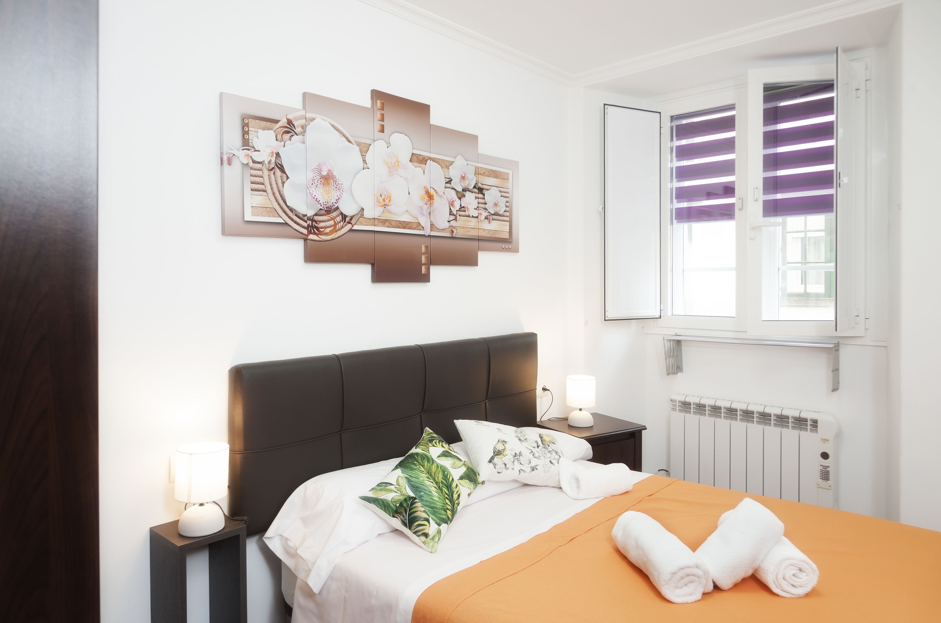 Quarto de hotel com uma cama, duas mesas de cabeceira com luminárias, um radiador e uma janela com persianas.
