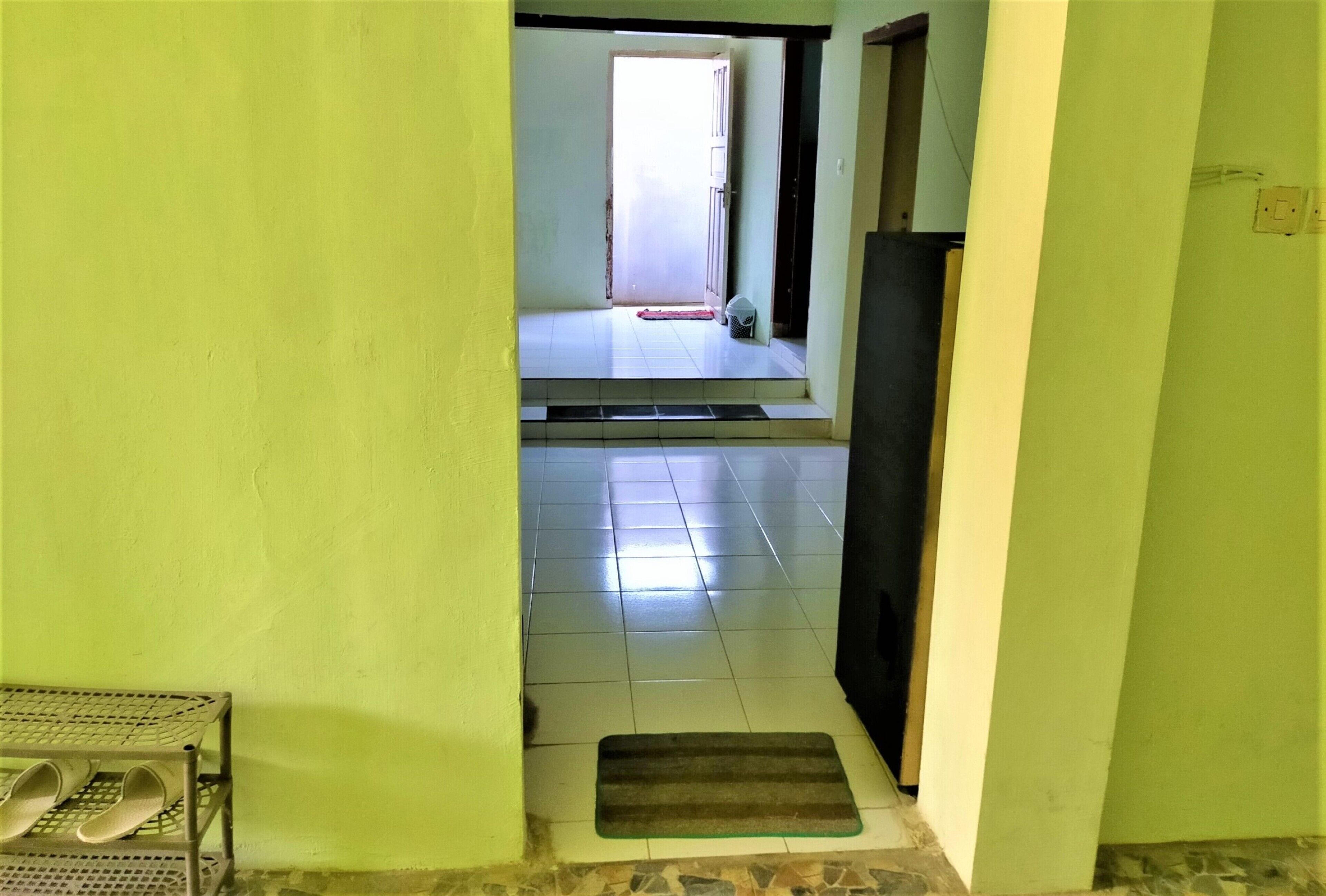 Corredor com um suporte de metal para sapatos, um tapete na entrada e uma porta que dá para um quarto com chão de azulejos.