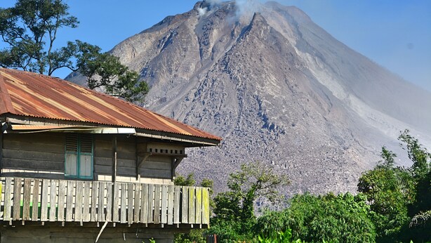 Uma casa de madeira rústica com telhado de metal corrugado, localizada em frente a uma montanha com uma coluna de fumaça visível, provavelmente um vulcão.