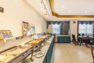 Buffet montado com várias estações de comida, área de refeições com mesas e cadeiras, e vista para o exterior através de uma janela.