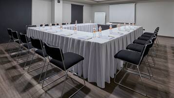 Uma sala de conferências com uma mesa comprida, cadeiras, garrafas de água e uma tela de projetor.