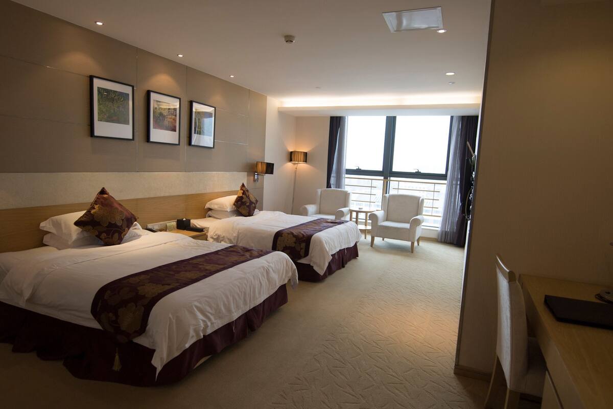 Quarto de hotel com duas camas, uma escrivaninha, uma cadeira e uma janela grande.