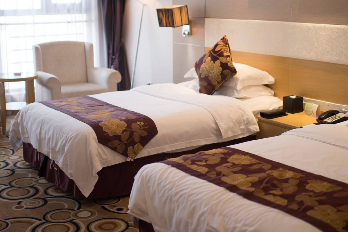 Quarto de hotel com duas camas, uma cadeira, uma escrivaninha e uma luminária.