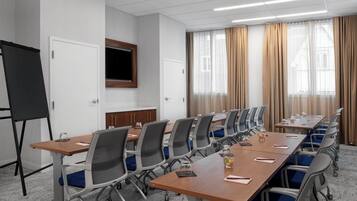 Sala de conferência com uma mesa comprida, várias cadeiras, tela de projetor e uma televisão na parede.