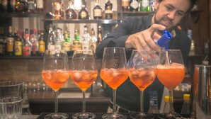 Um bartender preparando coquetéis num bar, com prateleiras cheias de garrafas de bebidas espirituosas ao fundo.