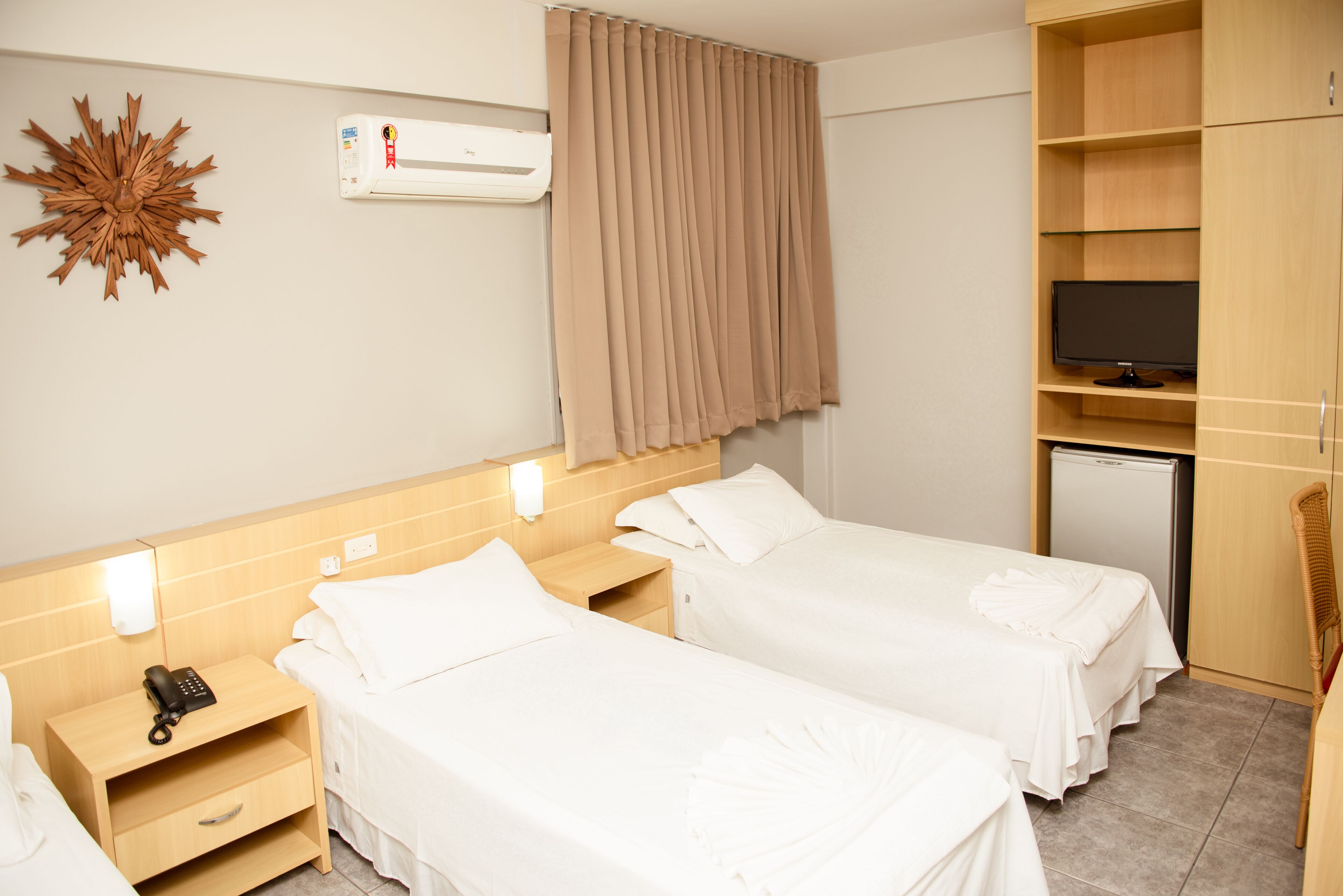 Quarto de hotel com duas camas de solteiro, uma escrivaninha com telefone, um pequeno frigorífico, uma televisão numa estante e um ar condicionado na parede.