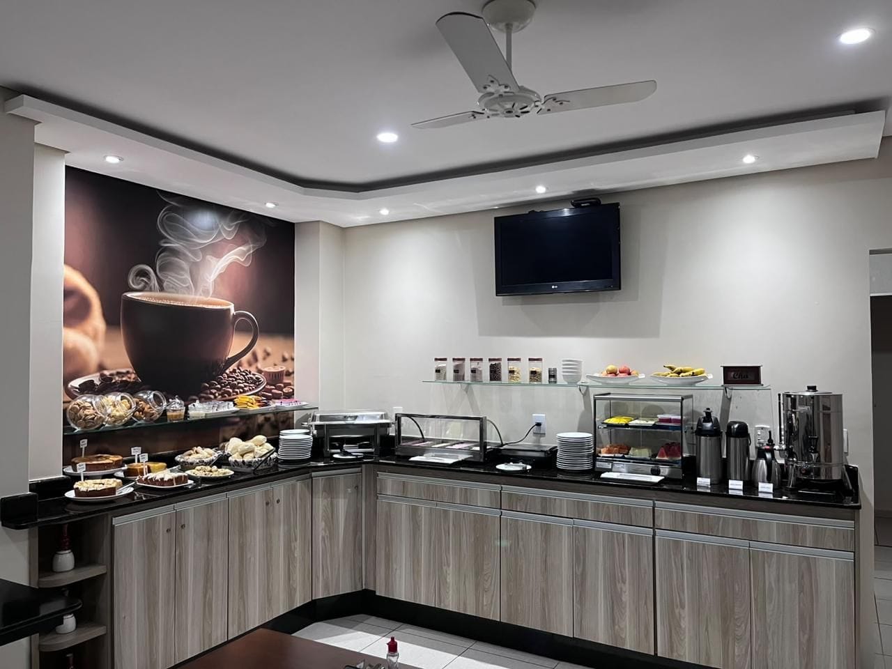 Estação de café moderna, com televisão de tela plana na parede, ventilador de teto e diversas opções de comida e bebida.