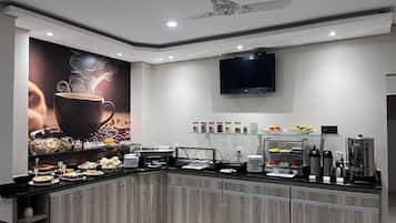 Estação de café moderna, com televisão de tela plana na parede, ventilador de teto e diversas opções de comida e bebida.