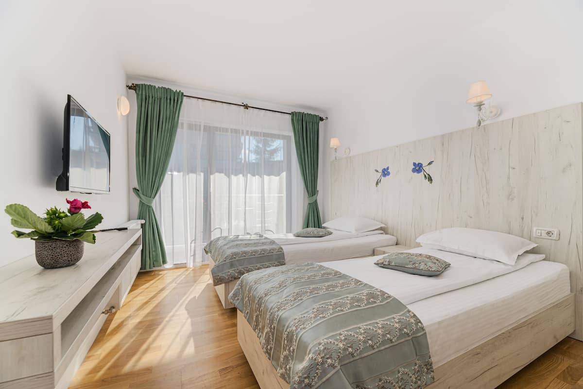 Quarto com duas camas, televisão, planta e janela com cortinas verdes.
