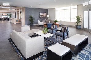 Uma sala de estar moderna com sofá, poltronas, mesa de centro e mesa de jantar. Há plantas em vasos e uma grande janela que proporciona iluminação natural.