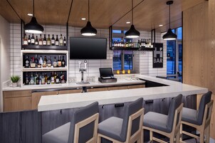 Uma área de bar com um balcão comprido, diversas garrafas de bebidas alcoólicas, uma televisão e banquetas.
