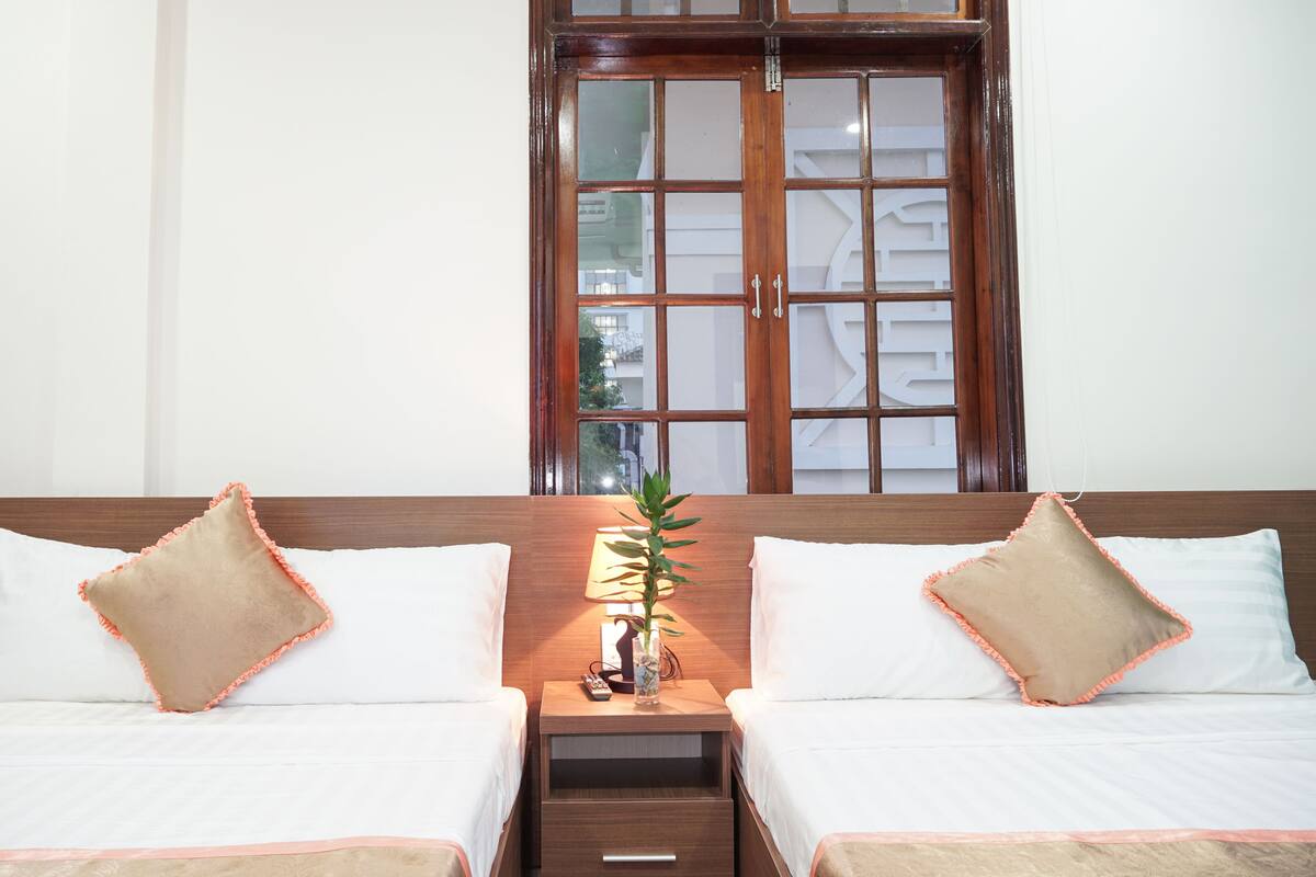 Quarto de hotel com duas camas, uma mesa de cabeceira com abajur e uma janela com vista para edifícios.