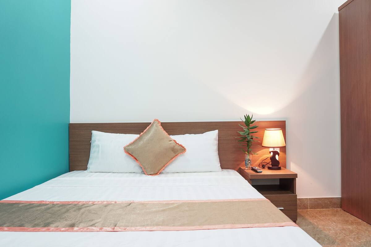 Uma cama bem arrumada com um edredom branco e bege, duas almofadas e uma almofada decorativa. Uma mesa de cabeceira com uma luminária e um controle remoto fica ao lado da cama.