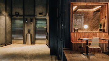 Elevador moderno com painéis digitais, balcão de recepção em madeira e área de estar com cadeira e mesa.