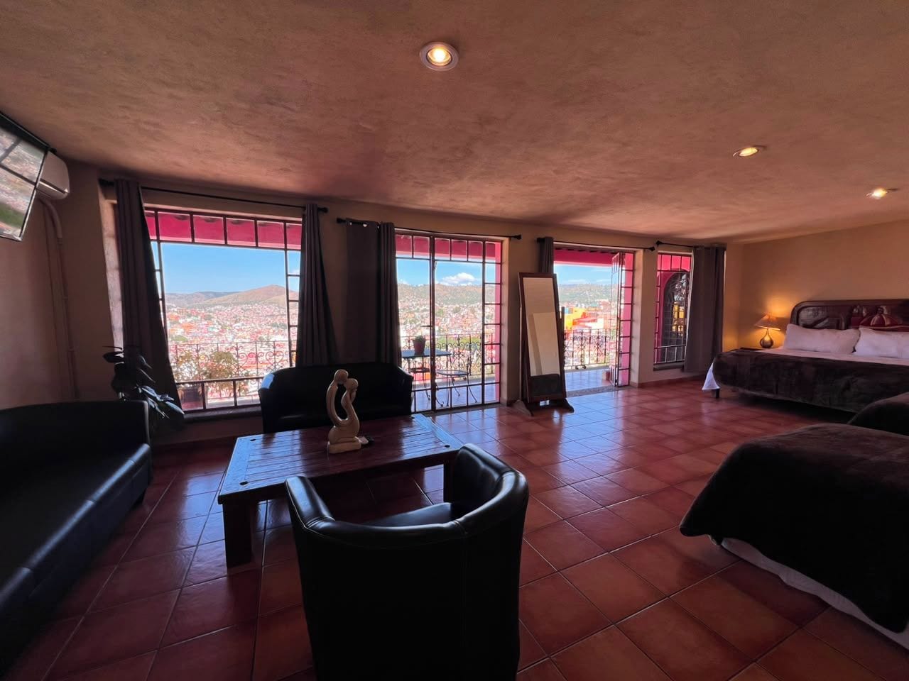 Quarto amplo com uma cama grande, um sofá, uma mesa e uma cadeira. Oferece vista para a paisagem urbana através de grandes janelas.