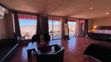 Quarto amplo com uma cama grande, um sofá, uma mesa e uma cadeira. Oferece vista para a paisagem urbana através de grandes janelas.