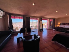 Quarto amplo com uma cama grande, um sofá, uma mesa e uma cadeira. Oferece vista para a paisagem urbana através de grandes janelas.