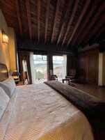 Quarto com cama grande, teto de madeira, vista da cidade e varanda.