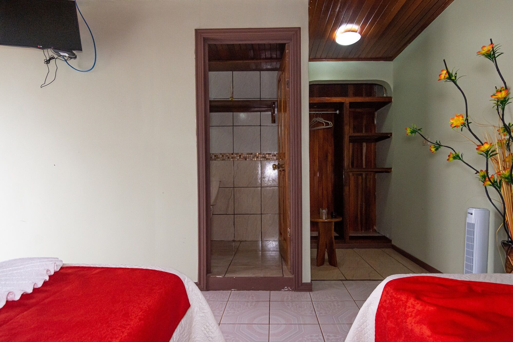Quarto de hotel com duas camas, porta de madeira, chão ladrilhado, uma mesinha e um vaso com flores.
