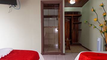 Quarto de hotel com duas camas, porta de madeira, chão ladrilhado, uma mesinha e um vaso com flores.
