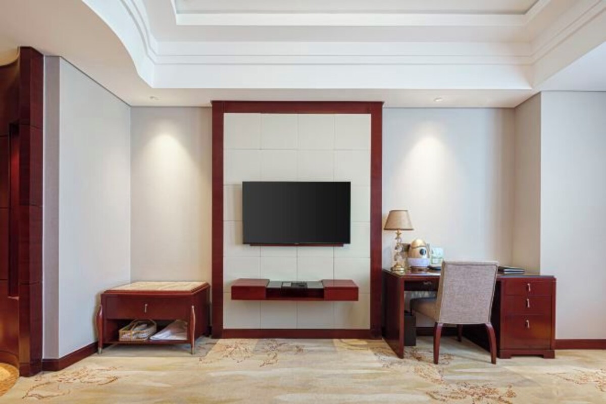 Quarto de hotel com televisão de tela plana fixada na parede, uma pequena escrivaninha com cadeira, luminária e um armário de madeira.