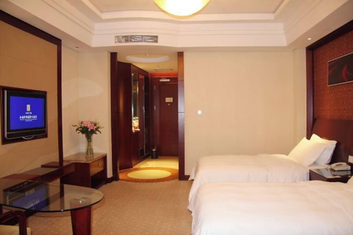 Quarto de hotel com televisão, duas camas, uma mesa de vidro, um vaso com flores e um corredor que dá para outro quarto.