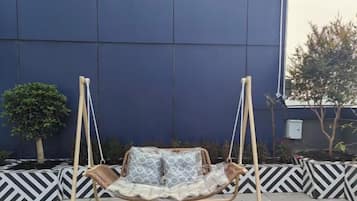 Uma cadeira de balanço de vime com almofadas em frente a uma parede azul, cercada por um canteiro geométrico.
