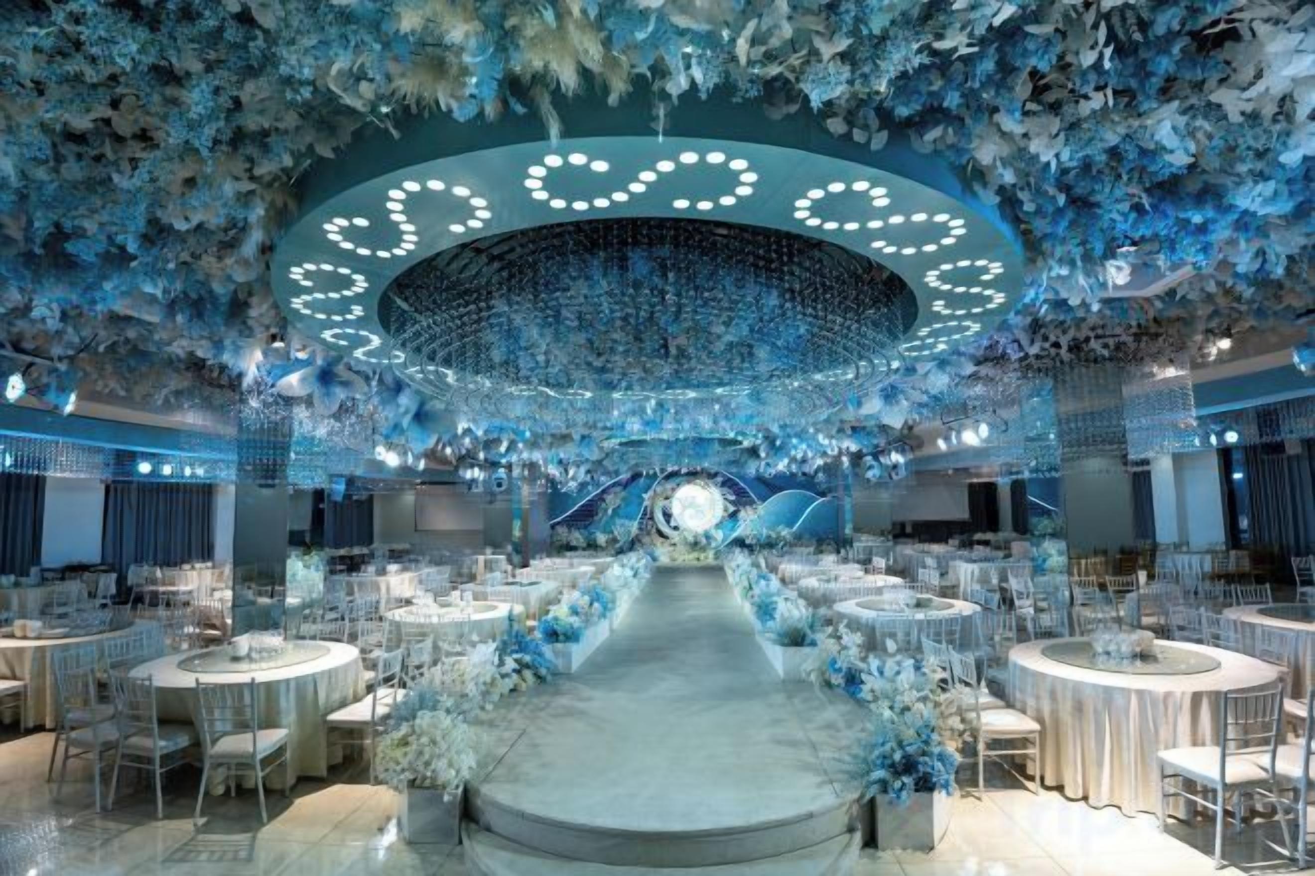 Espaçoso espaço interno com várias mesas redondas e cadeiras, um corredor central e um teto decorado com luzes pendentes e flores.